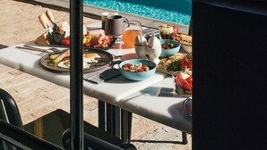Mesa de jantar com refeição posta à beira da piscina, com vista para uma piscina e um edifício de pedra.