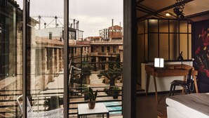 Quarto de hotel moderno com uma ampla parede de vidro que oferece vista para o pátio, uma pequena mesa de trabalho com cadeira, uma cama e um vaso de planta.