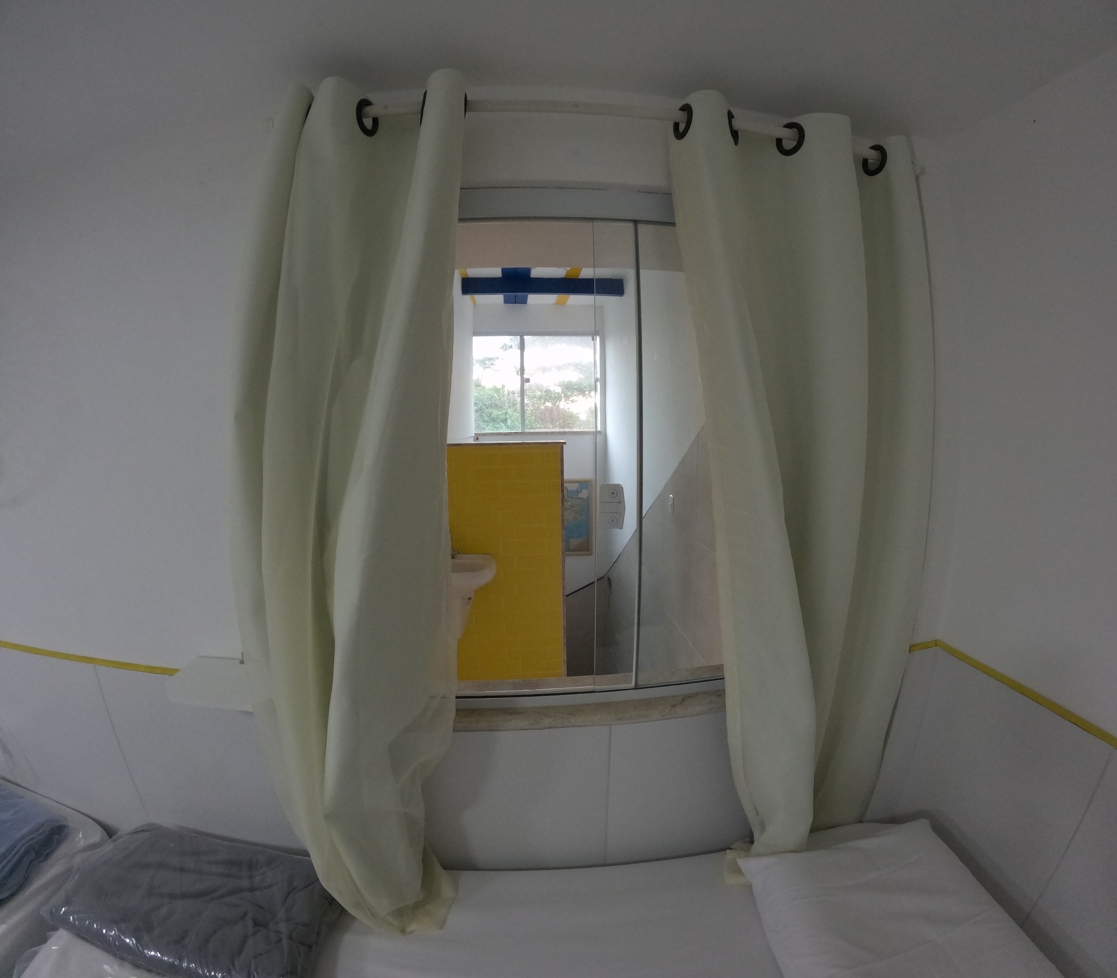 Quarto pequeno e estreito com janela, cortinas brancas, parede amarela e cama branca.