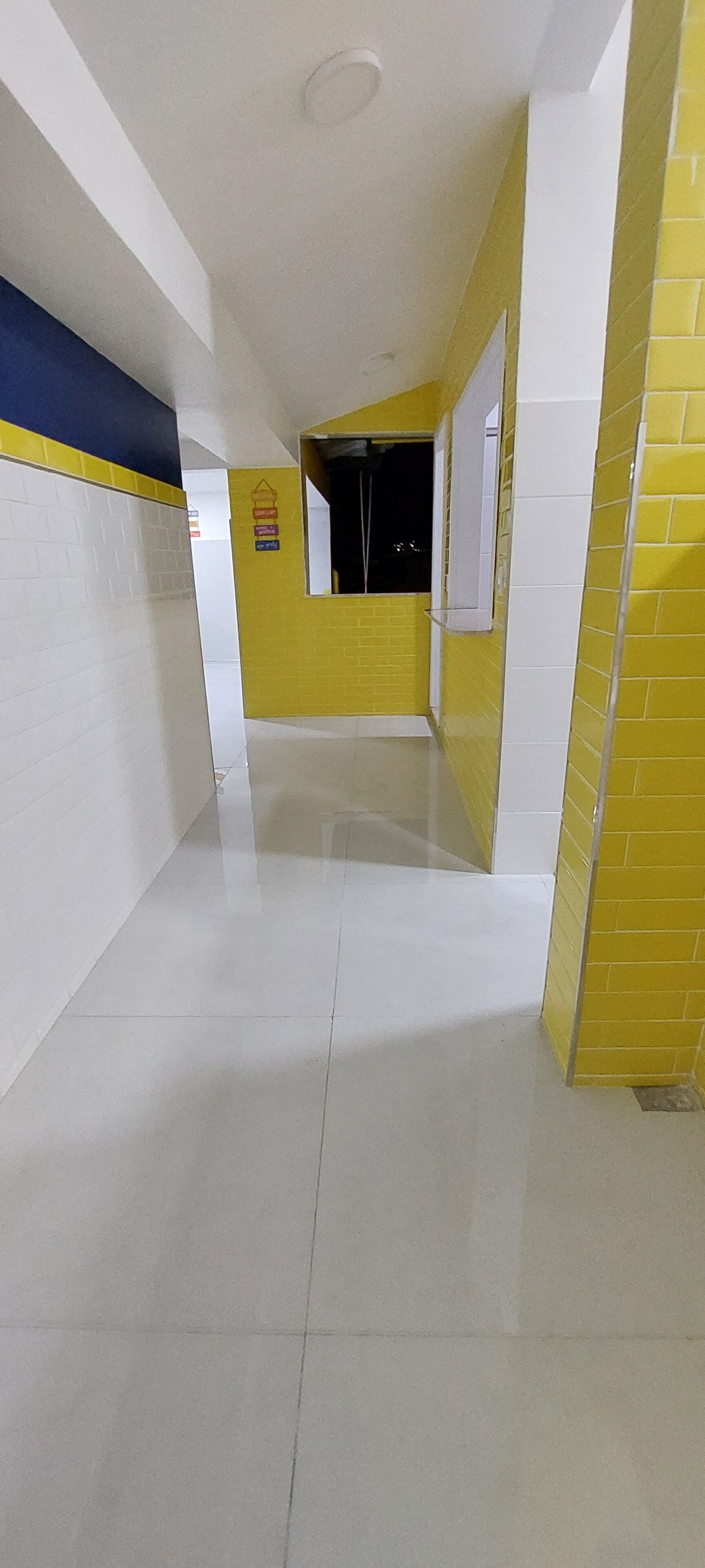 Um corredor com paredes amarelo e branco, piso de azulejos e uma janela no final.
