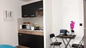 Cozinha compacta com armários pretos, micro-ondas e cafeteira. Há uma pequena área de jantar com duas cadeiras e uma mesa, e uma orquídea rosa em um vaso.