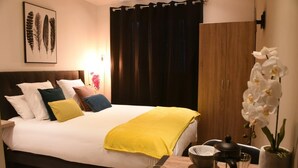 Cama bem arrumada com um xale amarelo, mesa de cabeceira com abajur e uma orquídea em vaso.