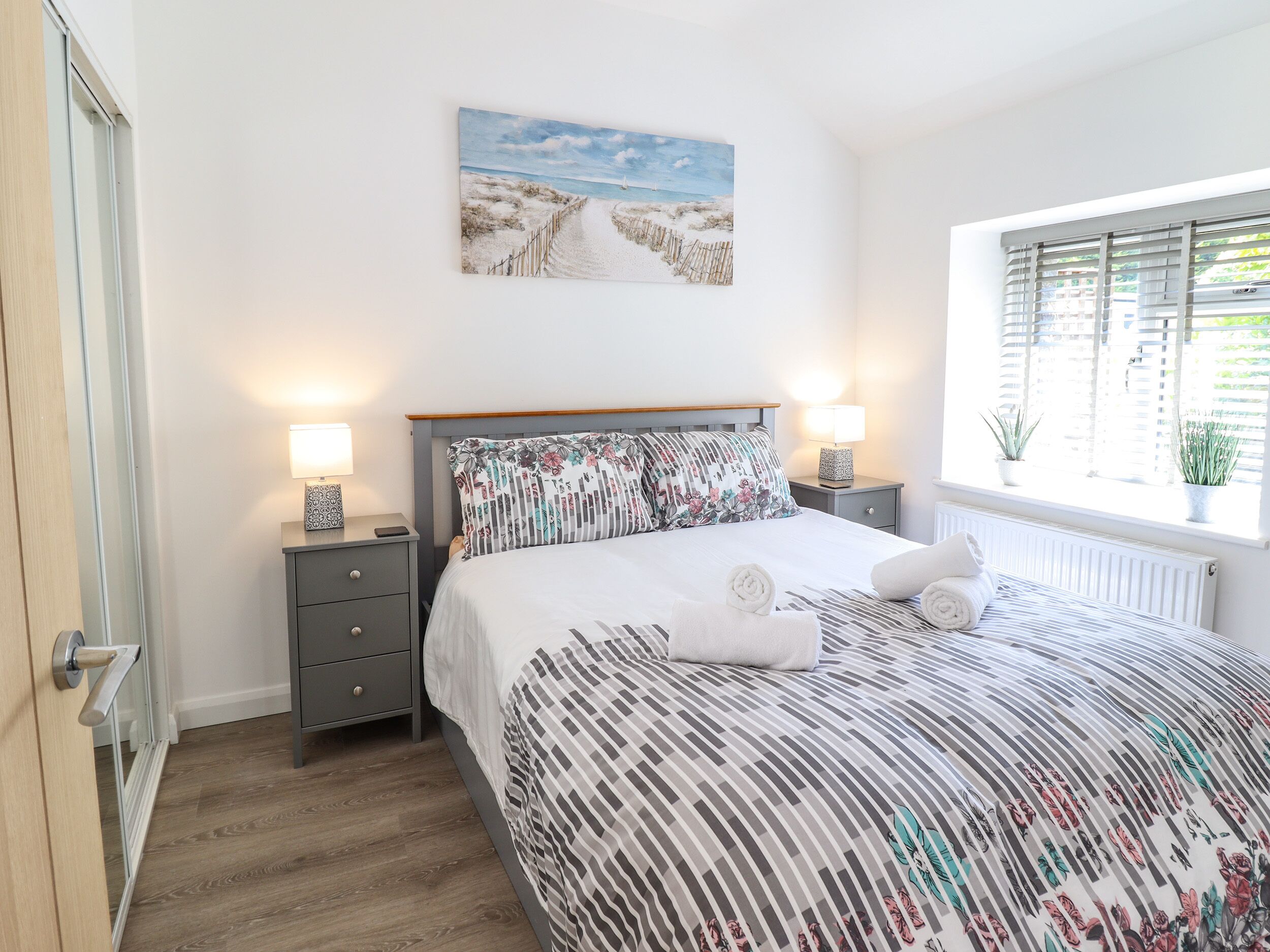Quarto com cama, mesas de cabeceira, um criado-mudo, janela com persianas e um quadro na parede.