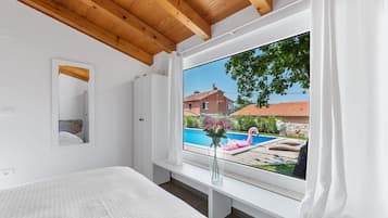 Quarto com uma grande janela, vigas de madeira no teto, uma cama, mesa de cabeceira e vista para uma piscina com boia de flamingo.