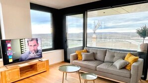 Uma sala de estar moderna com um sofá cinzento, duas mesinhas redondas, um móvel de entretenimento em madeira e uma janela ampla com vista para a cidade.