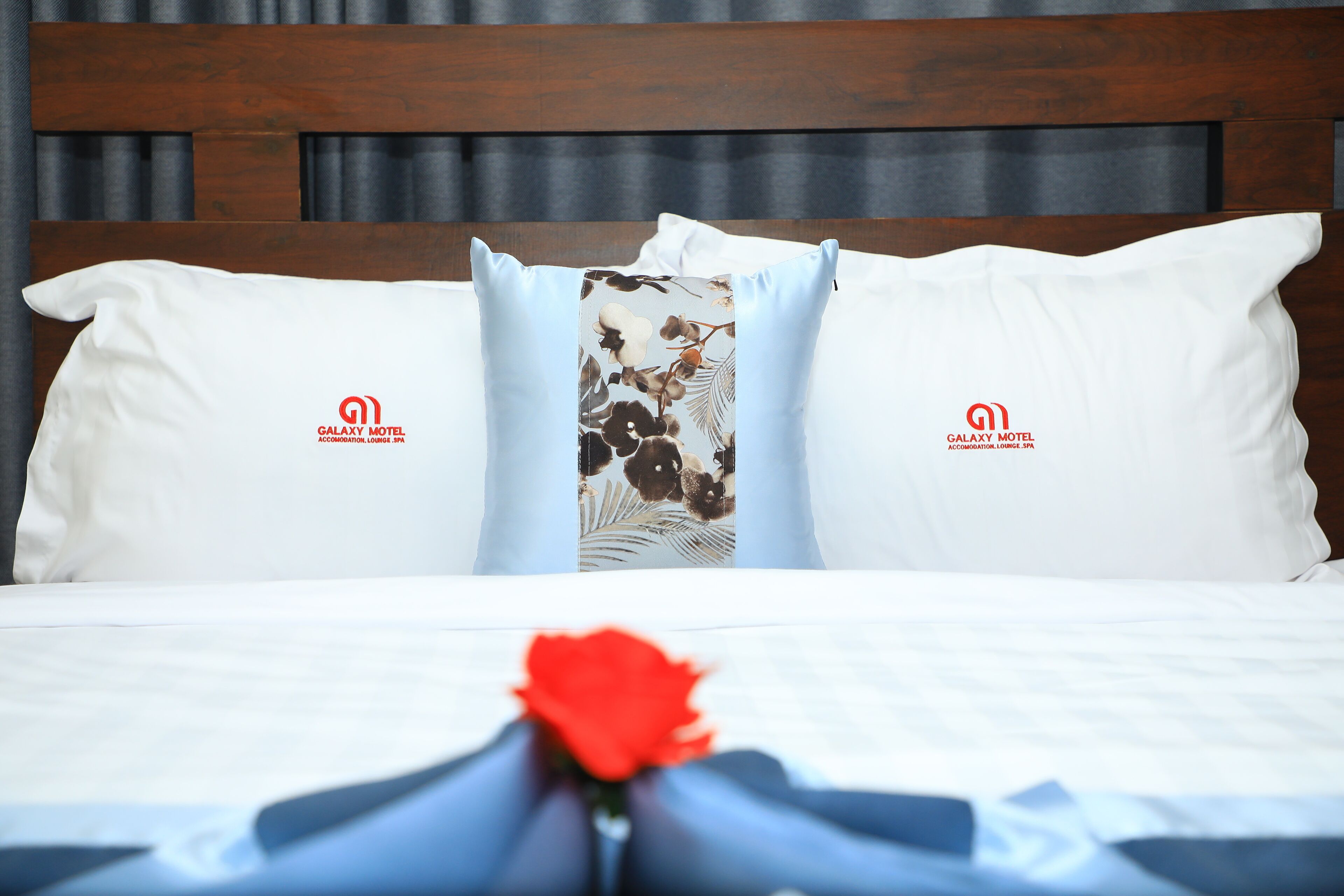 Cama bem arrumada com almofadas brancas e uma almofada floral, uma rosa vermelha e o logotipo do Galaxy Motel nas almofadas.