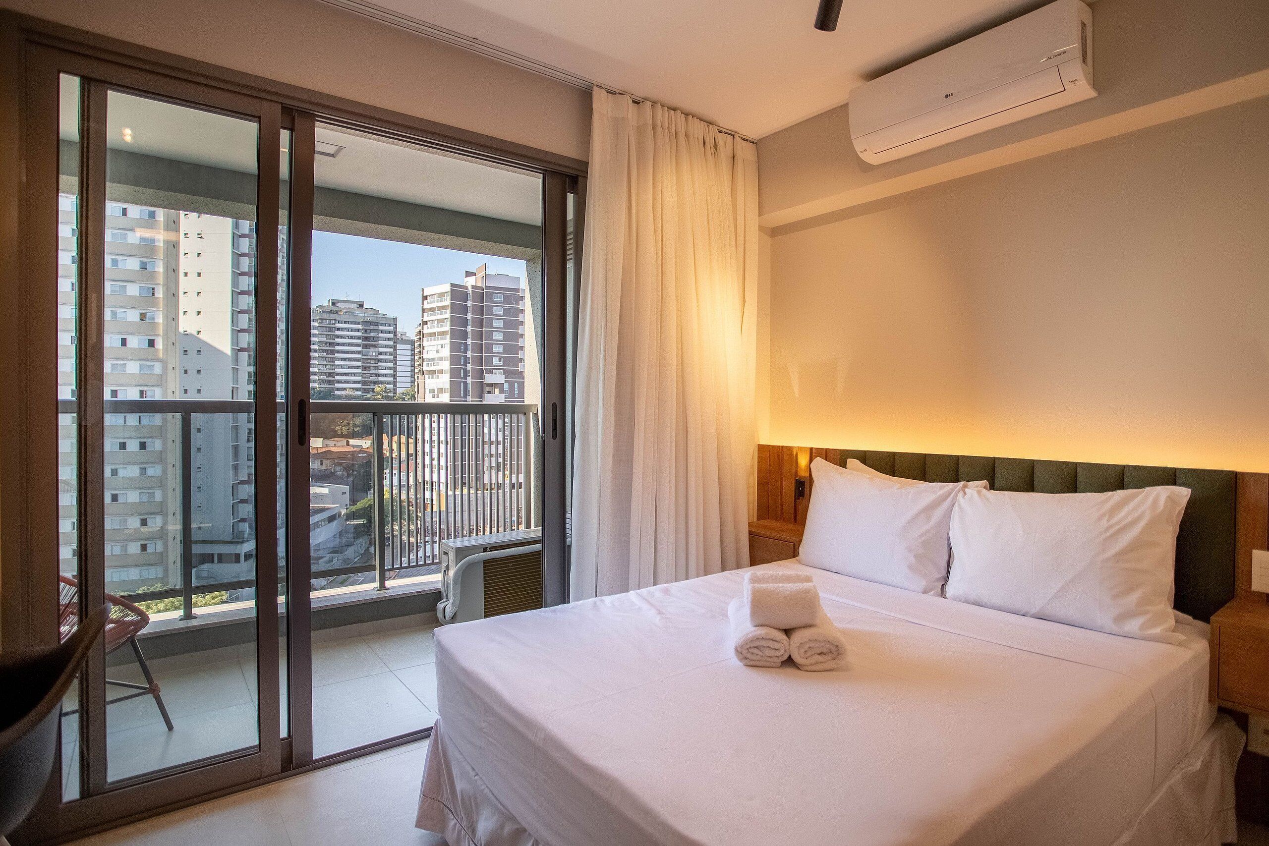 Quarto de hotel com cama, mesa de cabeceira, janela com cortinas e vista para edifícios.