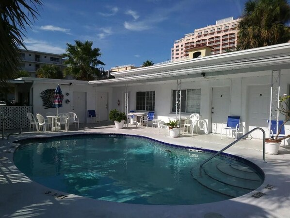 Piscina com espreguiçadeiras e mesas, rodeada por um edifício branco com várias portas e janelas.