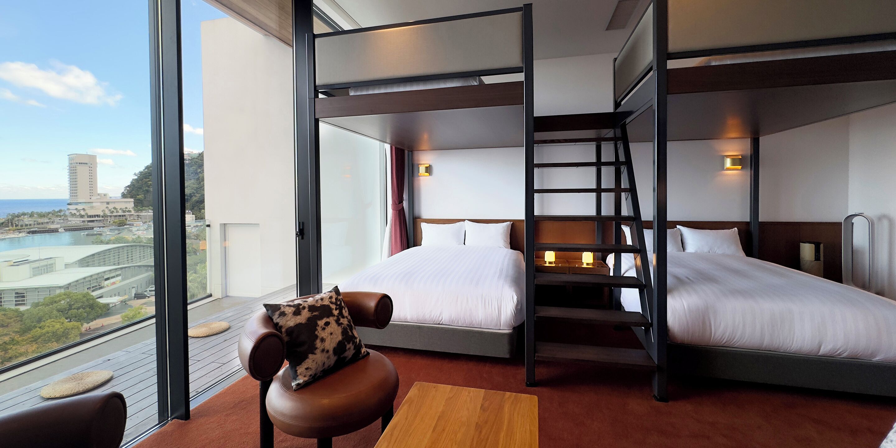 Quarto de hotel com varanda, duas camas, uma mesa de madeira e vista para a cidade e oceano.