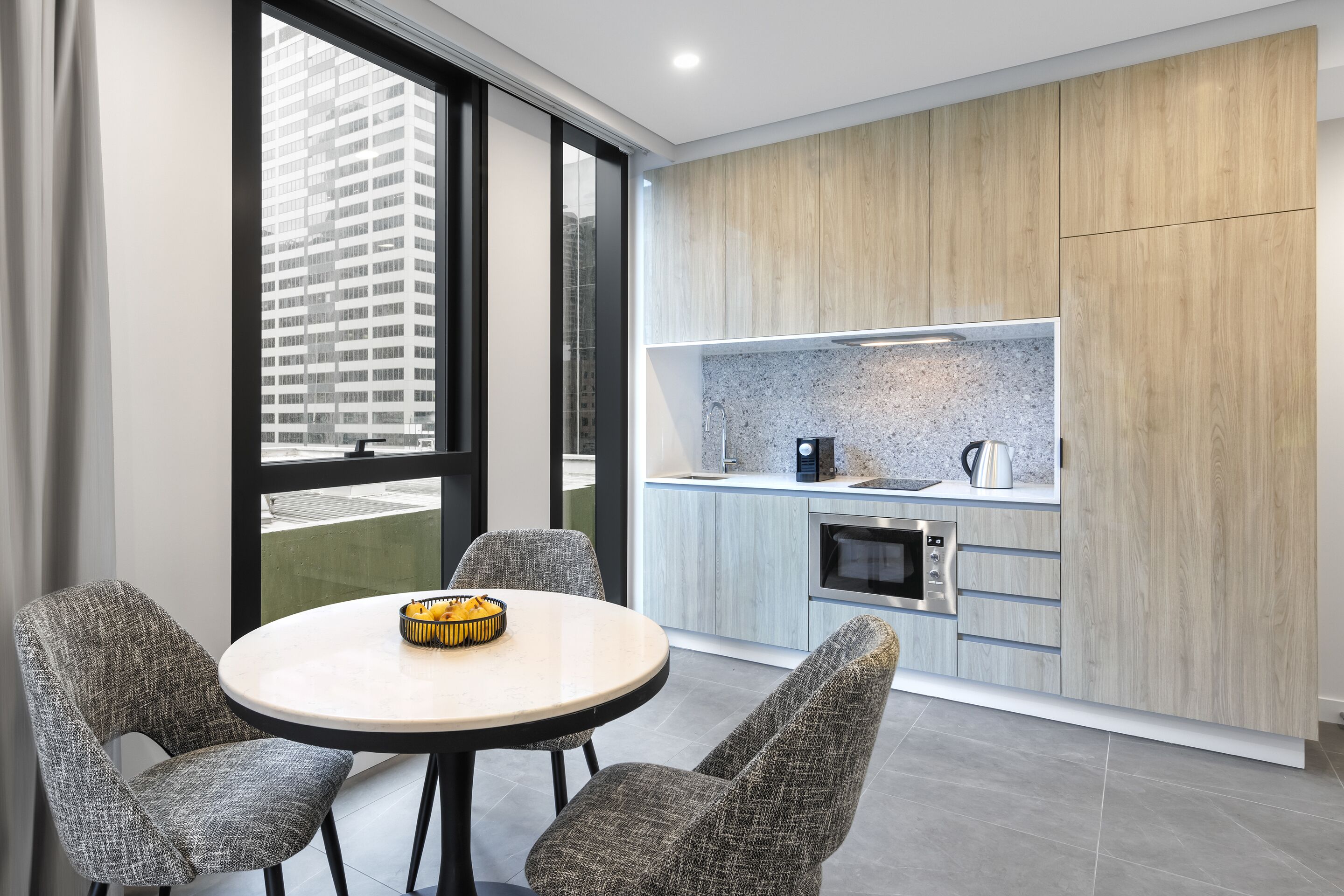 Uma cozinha moderna com forno embutido e micro-ondas, uma mesa de jantar redonda com quatro cadeiras e uma grande janela com vista para a paisagem urbana.
