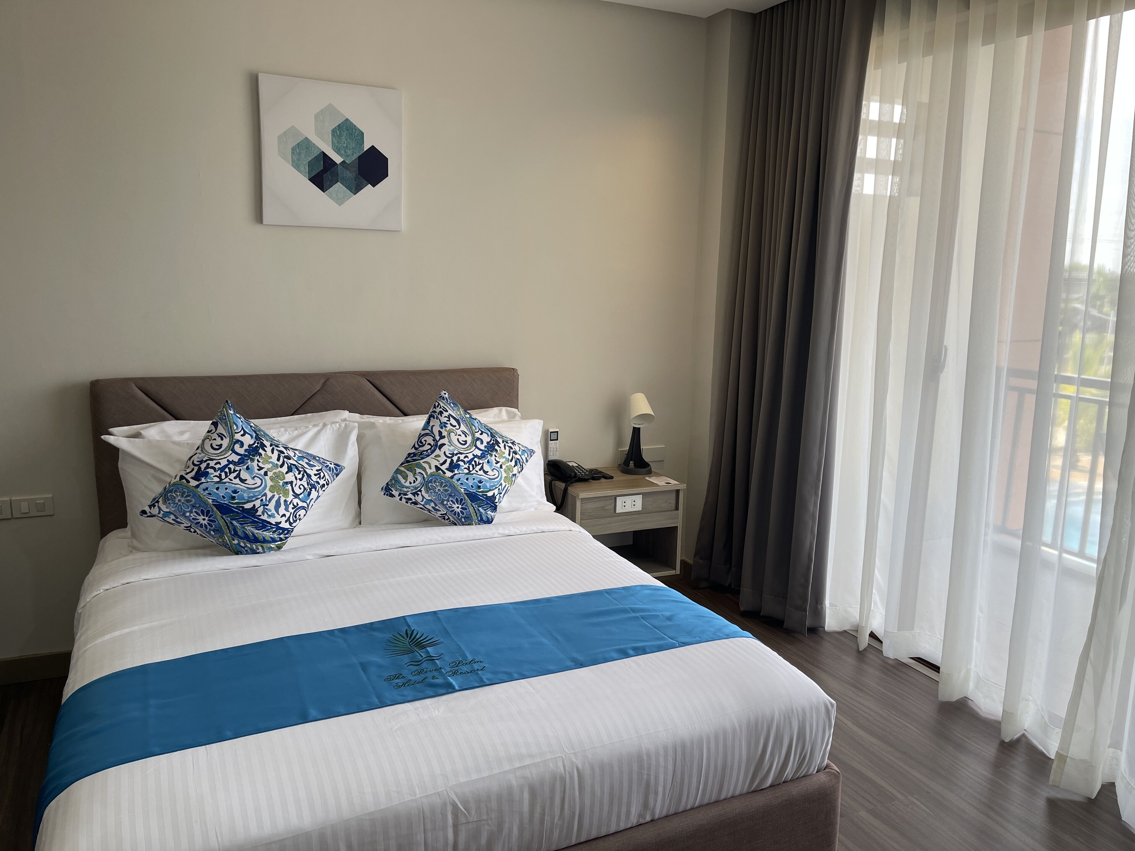 Cama bem arrumada com edredom azul e branco, duas almofadas decorativas, mesa de cabeceira com abajur e janela com cortinas leves.
