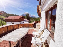 Um pátio com mesa de madeira e cadeiras brancas, com vista para uma cordilheira.