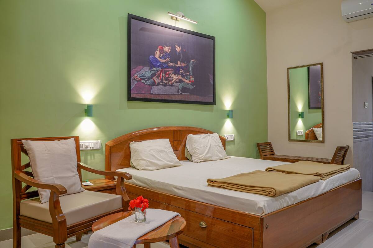 Quarto de hotel com uma cama grande, duas mesas de cabeceira, uma cadeira de madeira, uma pequena mesa com um vaso de flores e um quadro na parede.