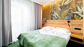 Quarto de hotel com uma cama, um cobertor verde, uma cabeceira de madeira, uma luminária fixada na parede e uma janela com cortinas leves.