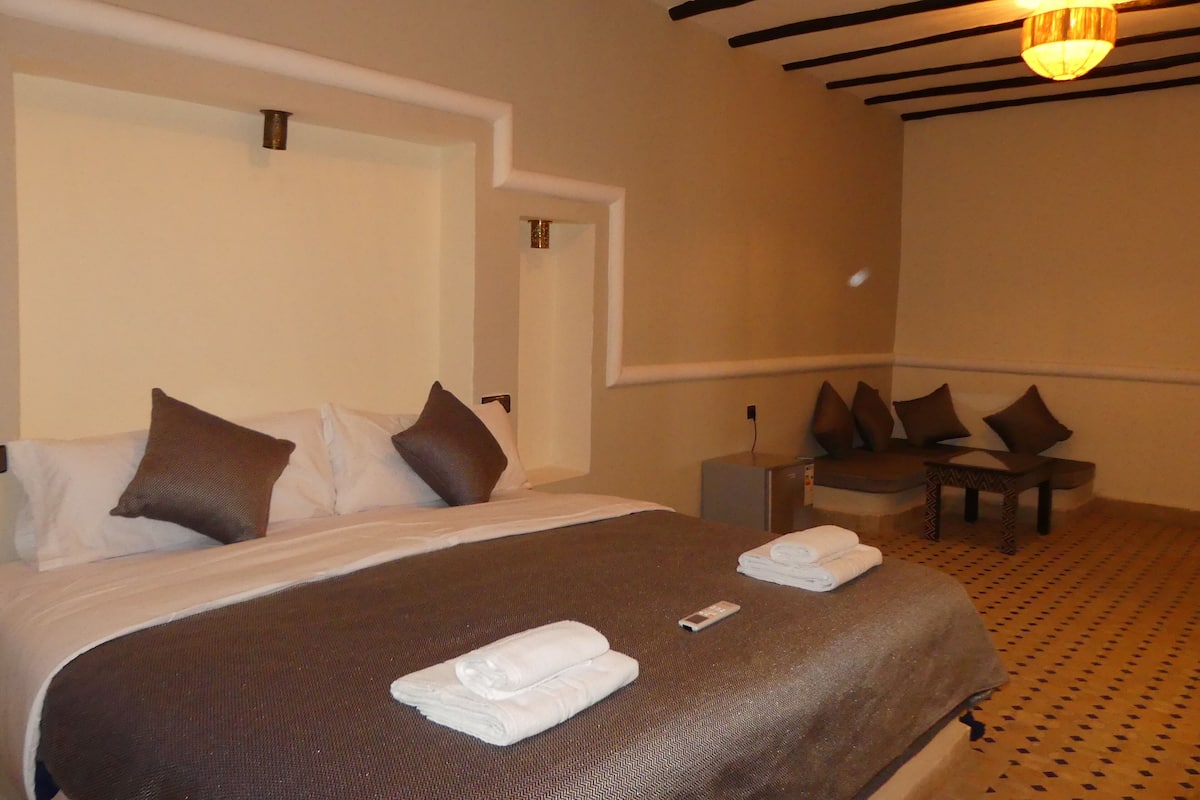 Quarto de hotel com cama, mesa de cabeceira, cadeira e uma pequena mesa com abajur.