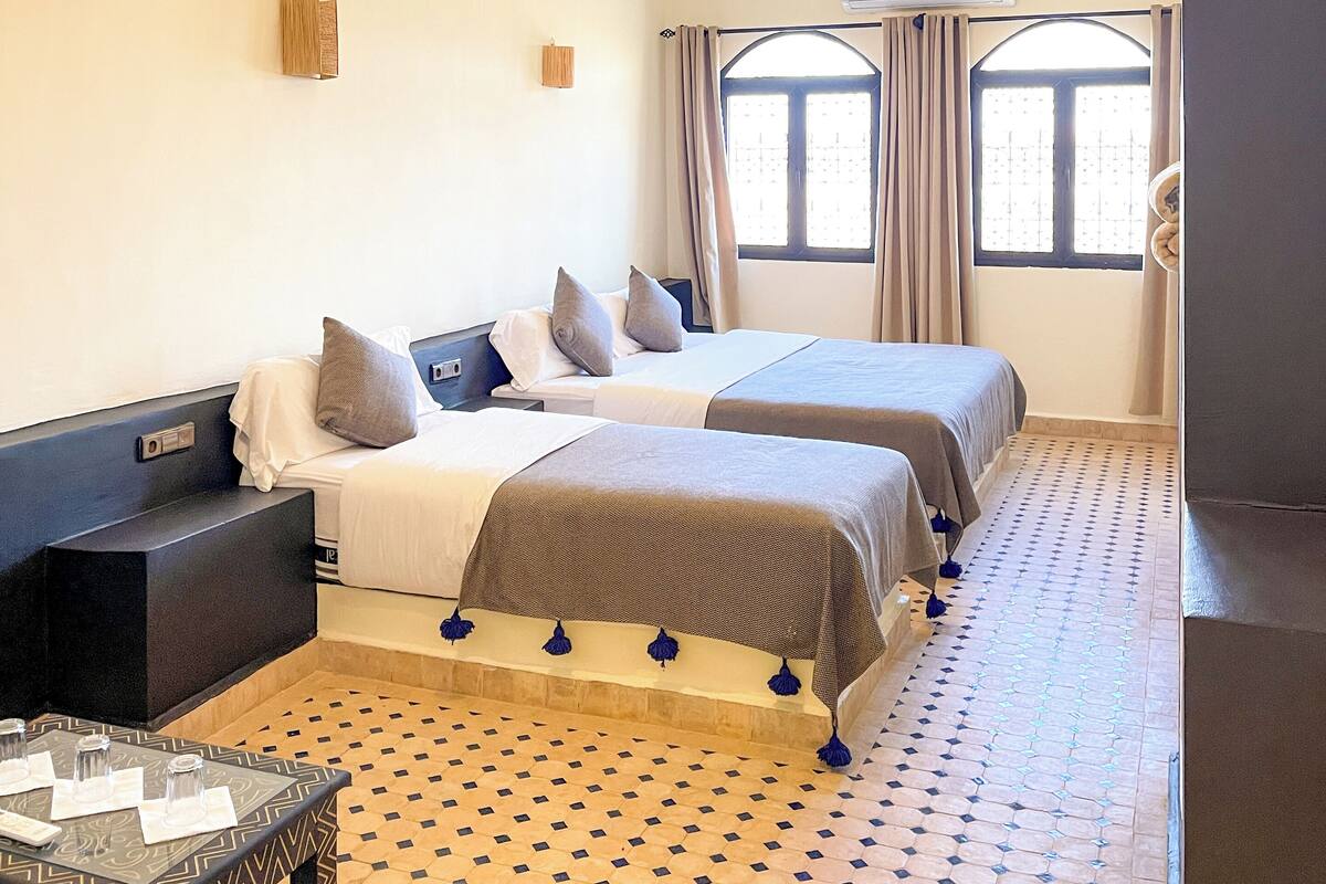 Quarto de hotel com duas camas, uma pequena mesa com copos, um sofá, ladrilhos no chão com padrão e teto com painéis de madeira e luminárias pendentes.