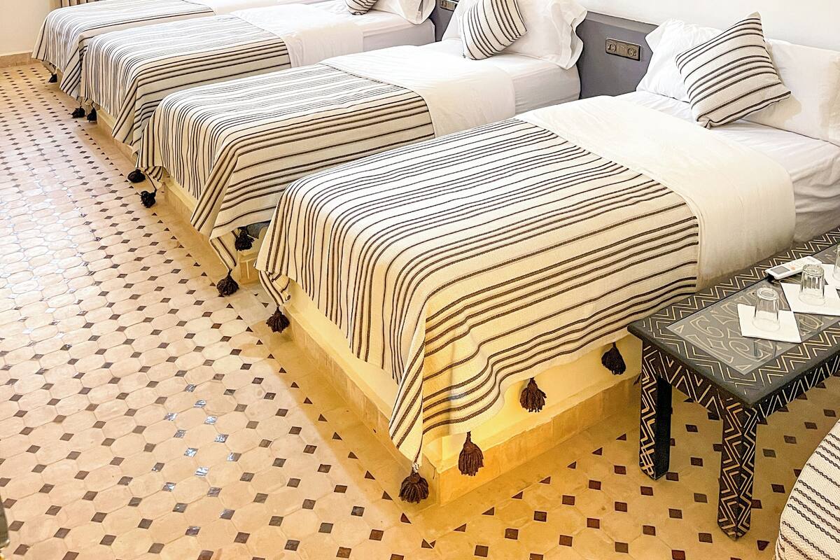Quarto de hotel com três camas, piso estampado, lustre e detalhes decorativos na parede.