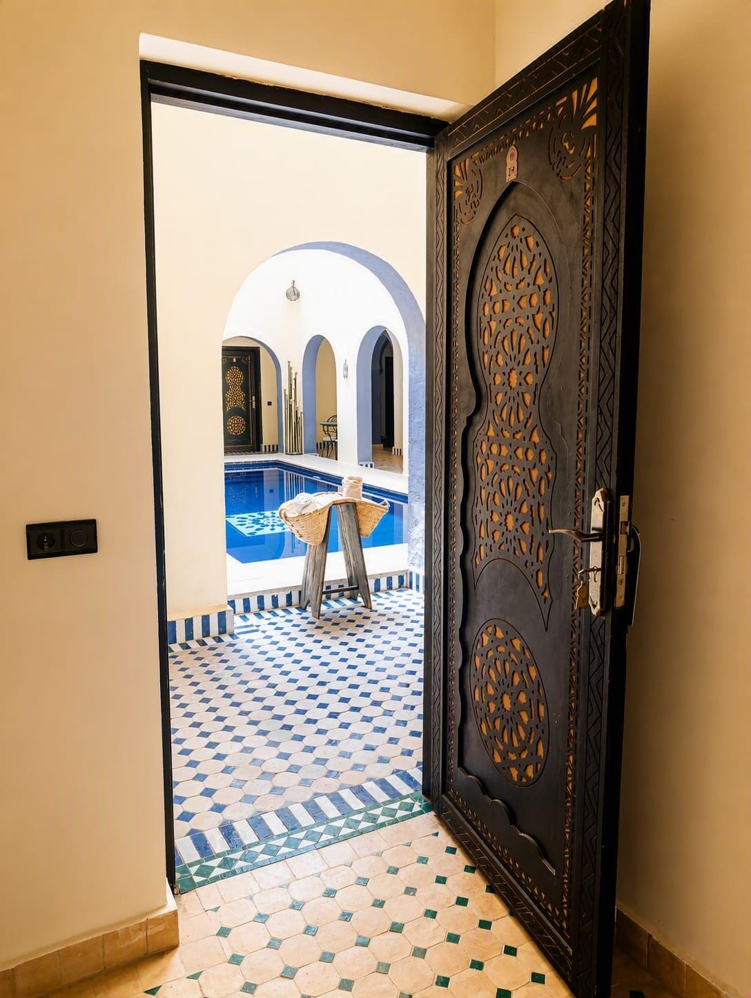 Uma porta aberta que dá para uma área de piscina com piso de azulejos e uma porta decorativa com padrões elaborados.