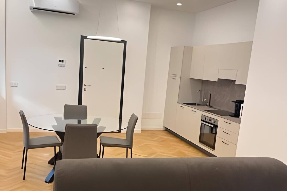 Uma sala de estar moderna com sofá, uma mesa de jantar com cadeiras, uma área de cozinha com armários e eletrodomésticos, e uma porta.