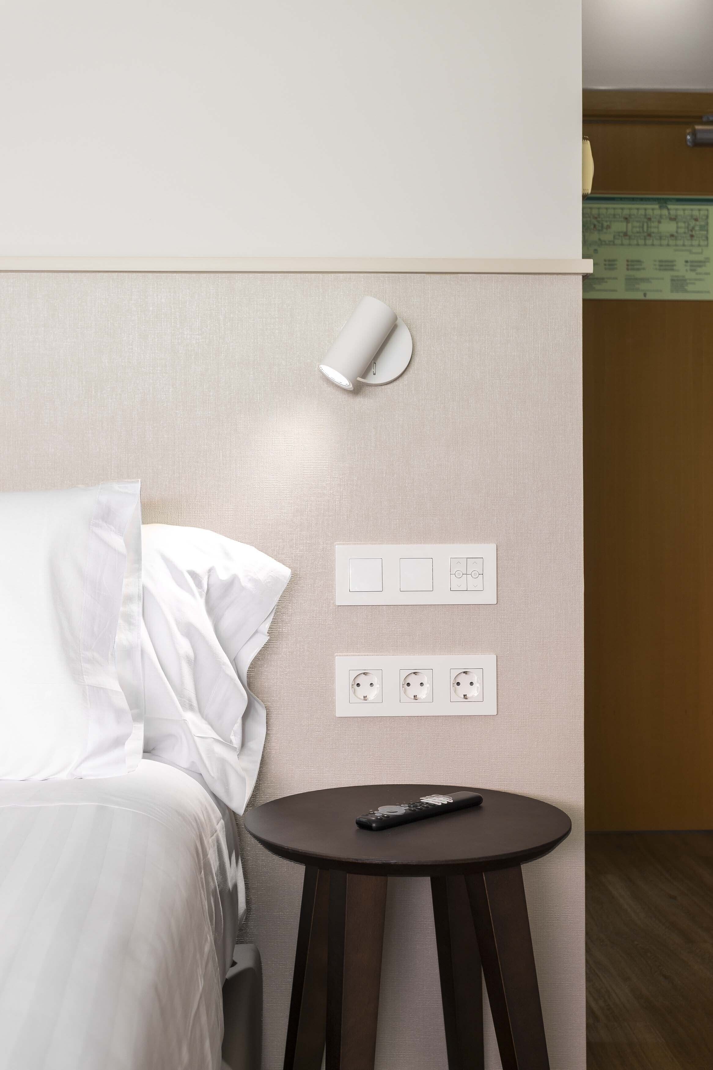 Quarto de hotel com cama, mesa de cabeceira com controle remoto e tomadas na parede.