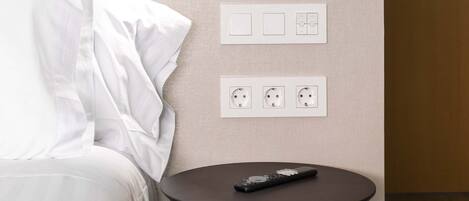 Quarto de hotel com cama, mesa de cabeceira com controle remoto e tomadas na parede.