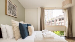 Quarto de hotel com cama grande, várias almofadas, vista para edifícios e janela com cortinas.
