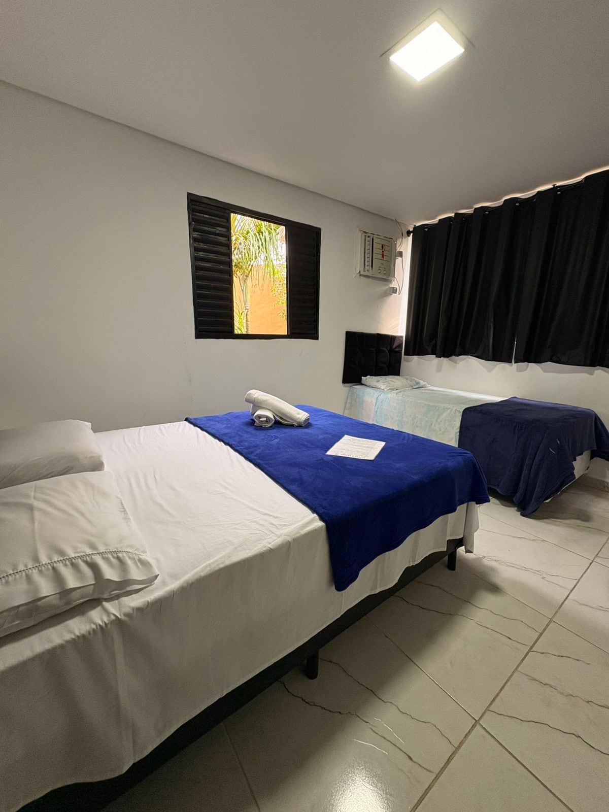Quarto de hotel com duas camas, janela com persianas, ar condicionado e luminária no teto.