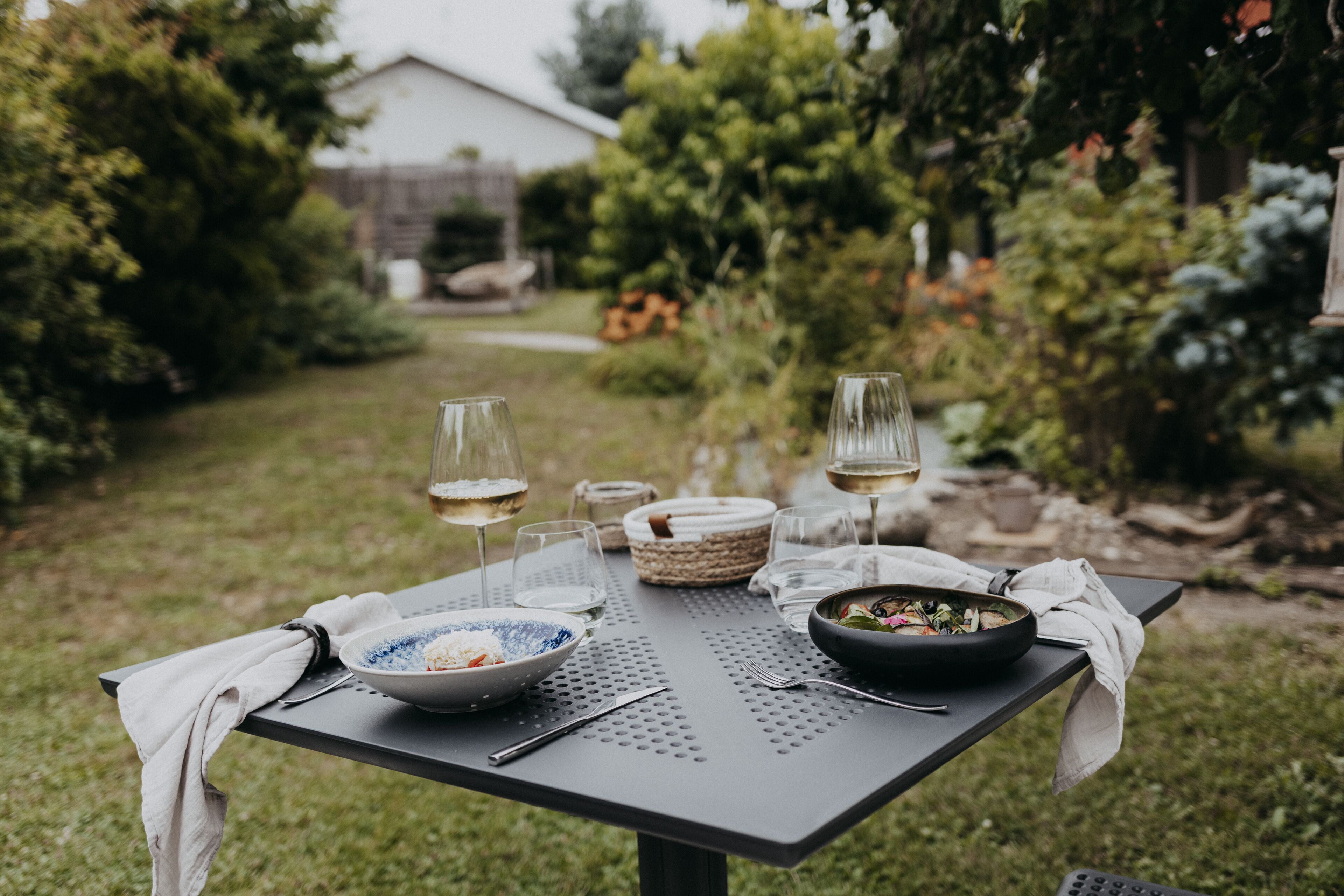 Mesa de jantar ao ar livre posta com pratos, copos e talheres, cercada por vegetação e uma casa ao fundo.