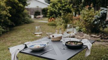 Mesa de jantar ao ar livre posta com pratos, copos e talheres, cercada por vegetação e uma casa ao fundo.