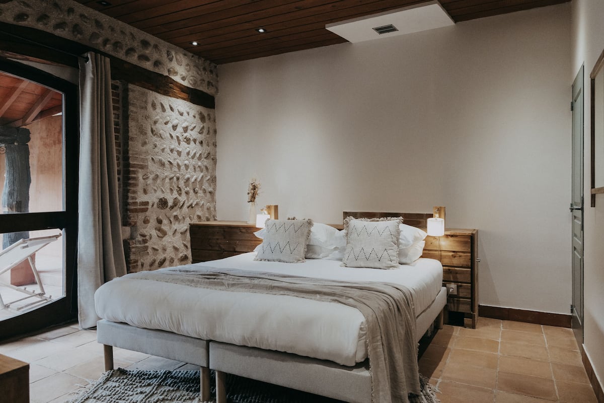 Um quarto com cama, mesas de cabeceira, luminária, janela com cortinas e parede de pedra.