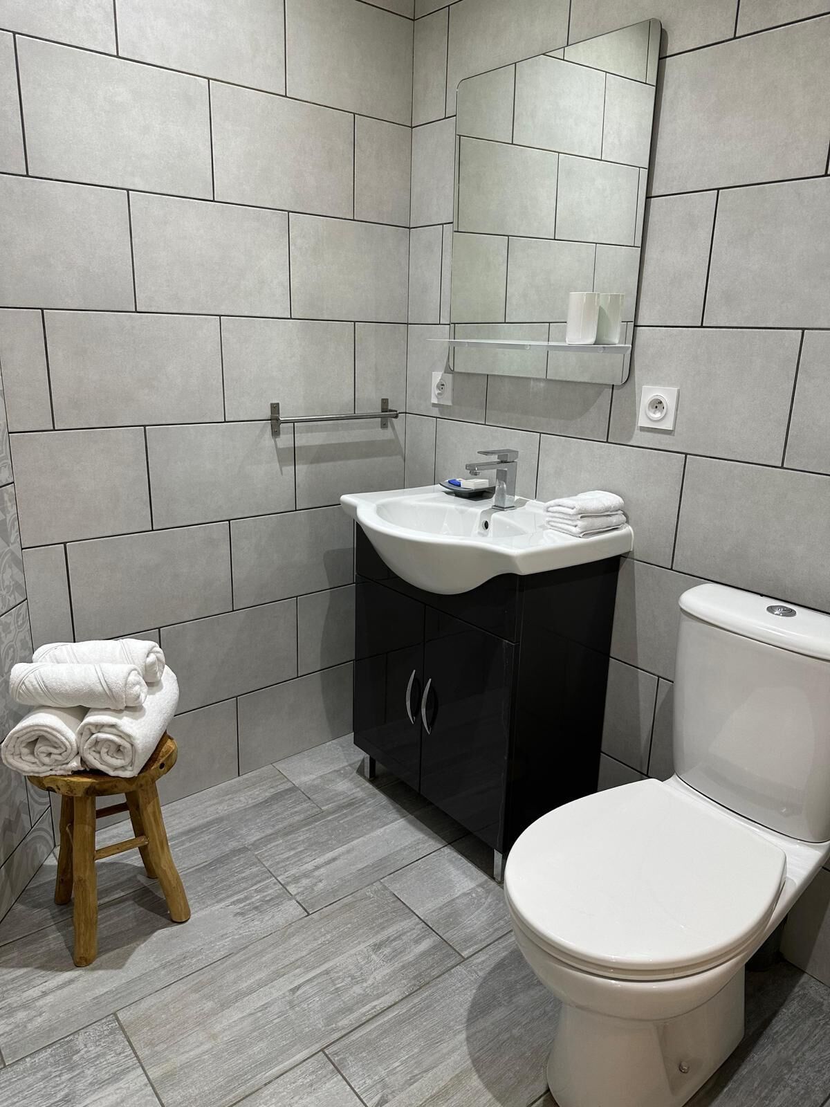 Casa de banho moderna com azulejos cinzentos, uma sanita, um lavatório branco, um armário preto e um banquinho de madeira com toalhas.