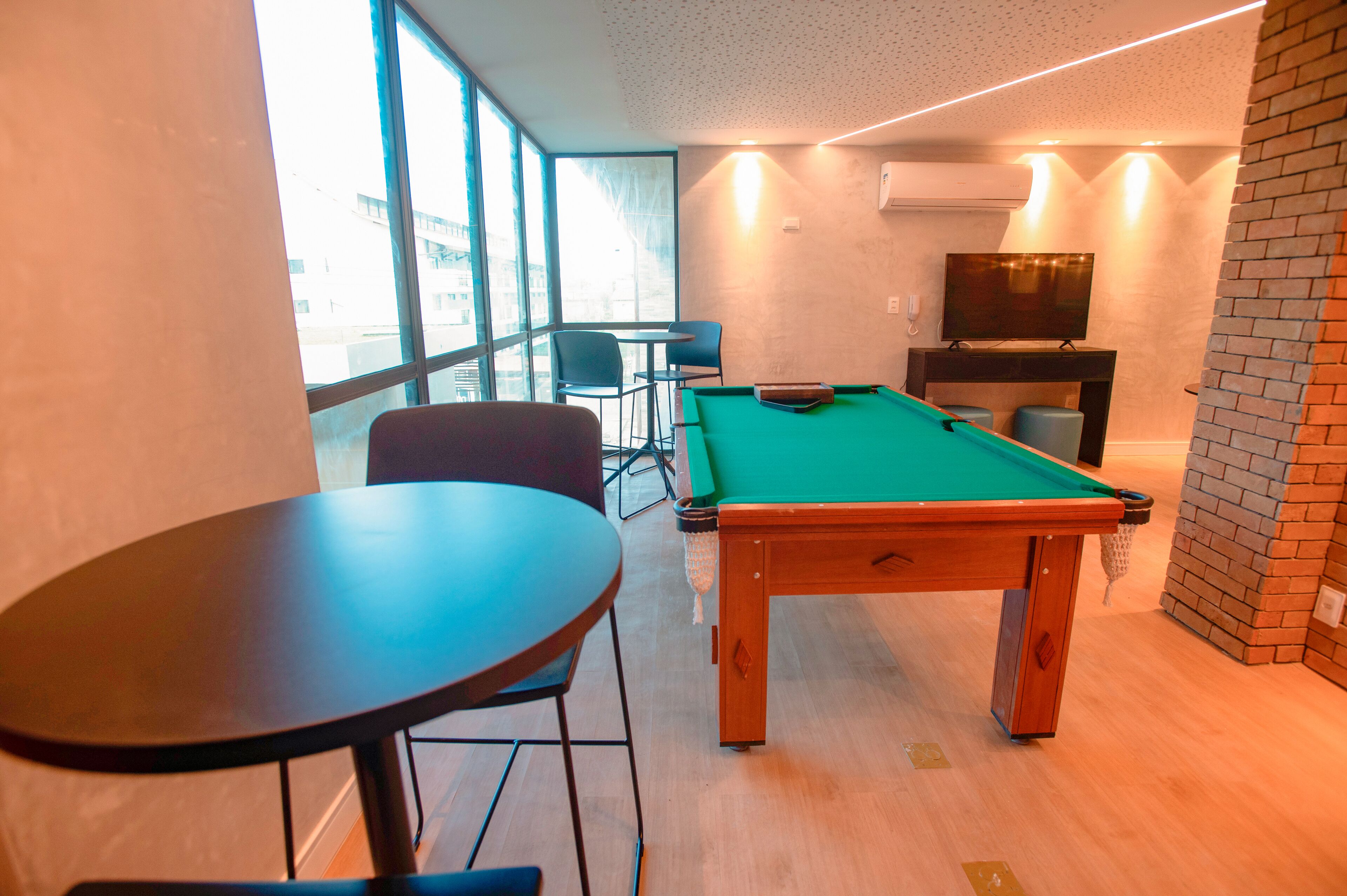 Mesa de bilhar com cadeiras, televisão e parede de tijolos num espaço com amplas janelas.