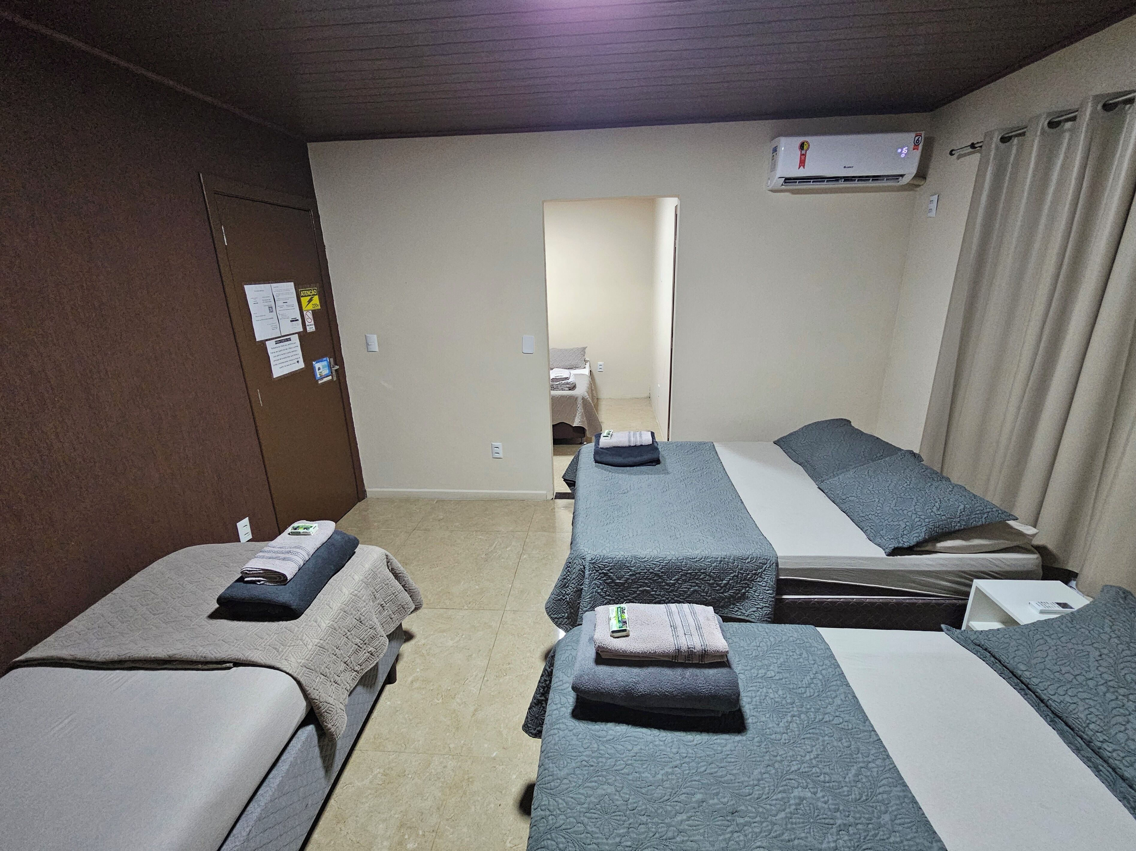 Quarto de hotel com duas camas de solteiro, uma pequena mesa, uma cadeira, um ar condicionado e uma porta com um quadro de avisos.