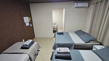 Quarto de hotel com duas camas de solteiro, uma pequena mesa, uma cadeira, um ar condicionado e uma porta com um quadro de avisos.