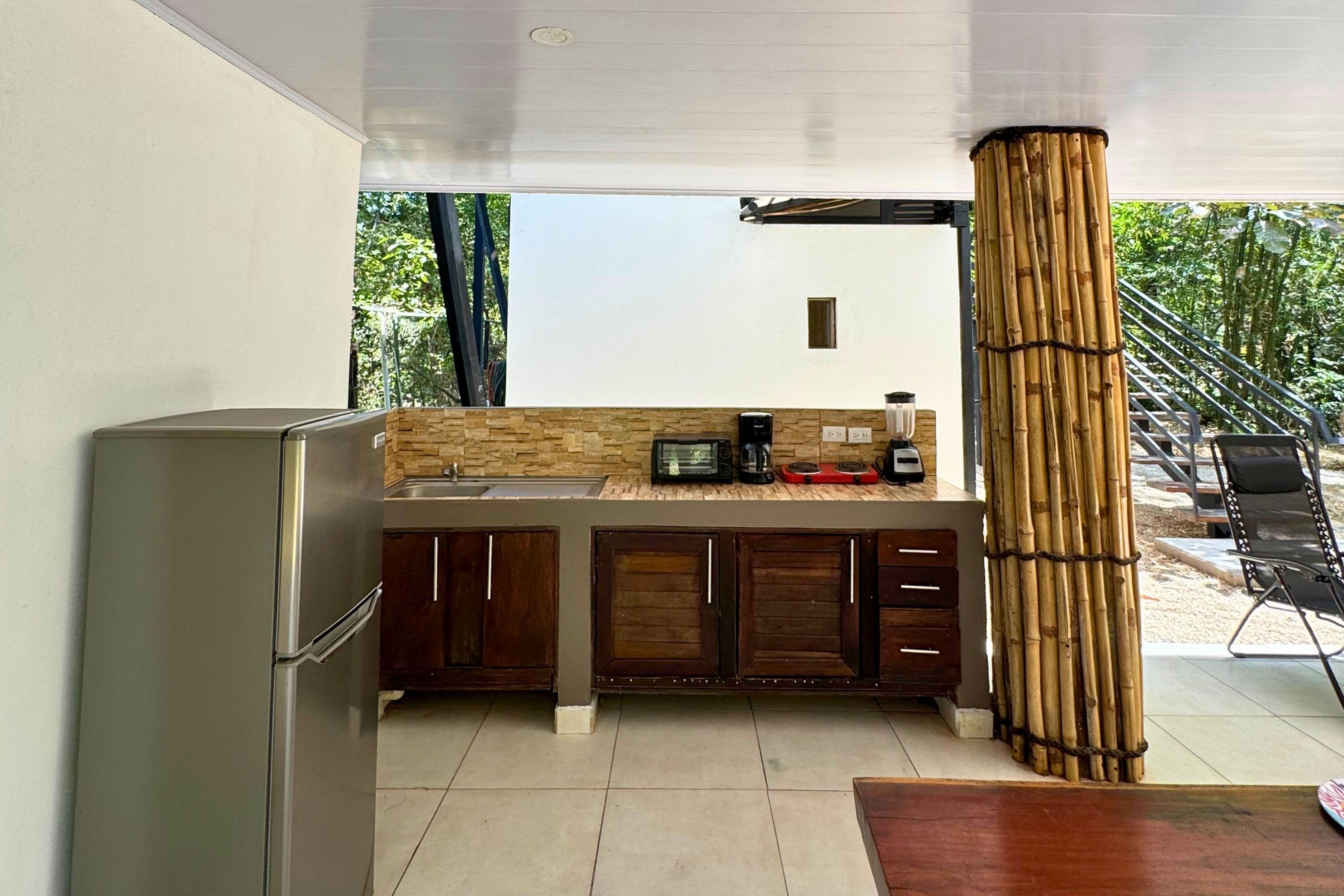 Cozinha compacta com geladeira, armários de madeira, painel de backsplash de pedra e um pilar de bambu.