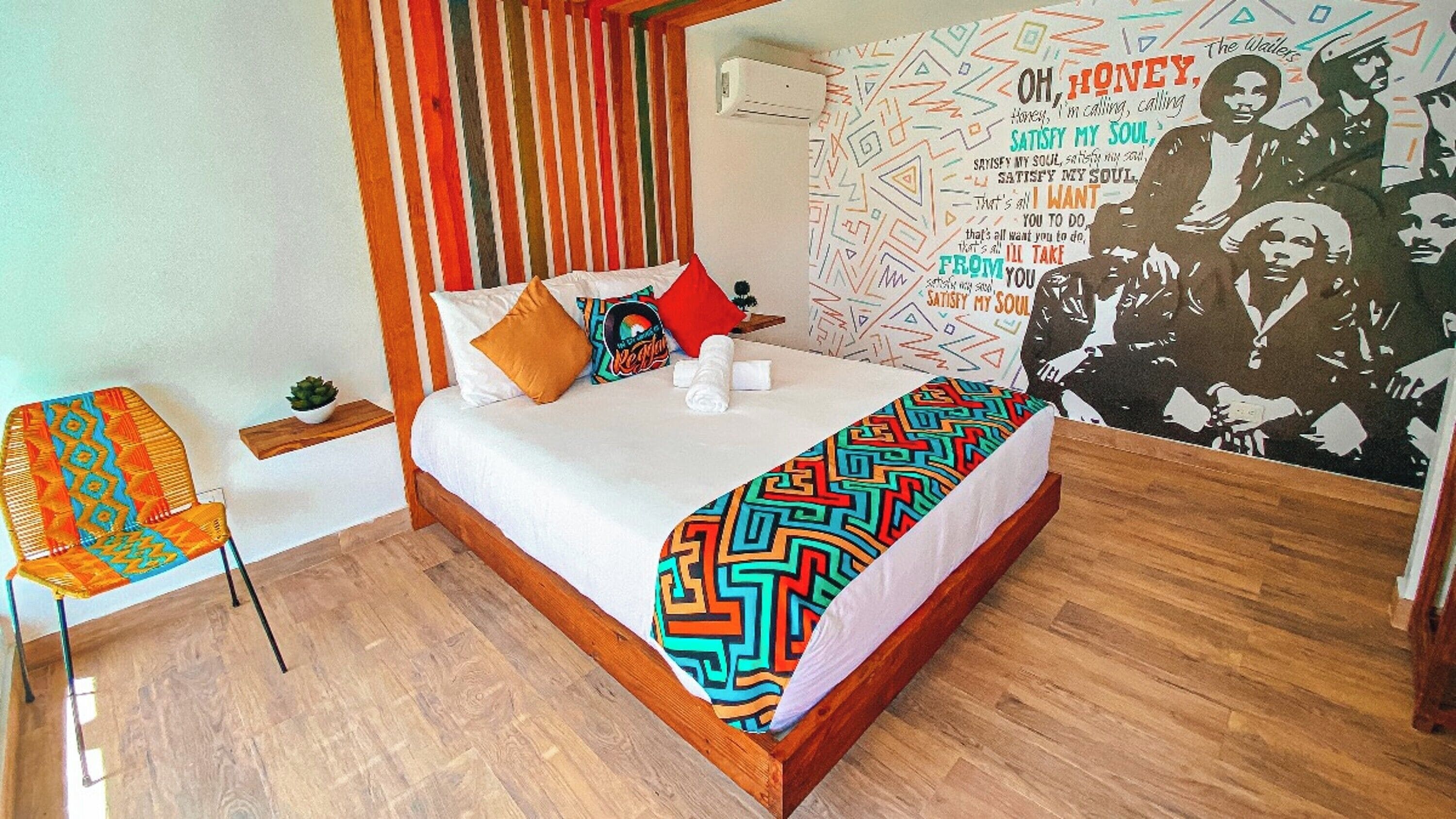 Um quarto com uma cama de madeira com dossel, um edredom colorido com padrão, uma pequena mesa de cabeceira com uma planta e uma cadeira com design geométrico.