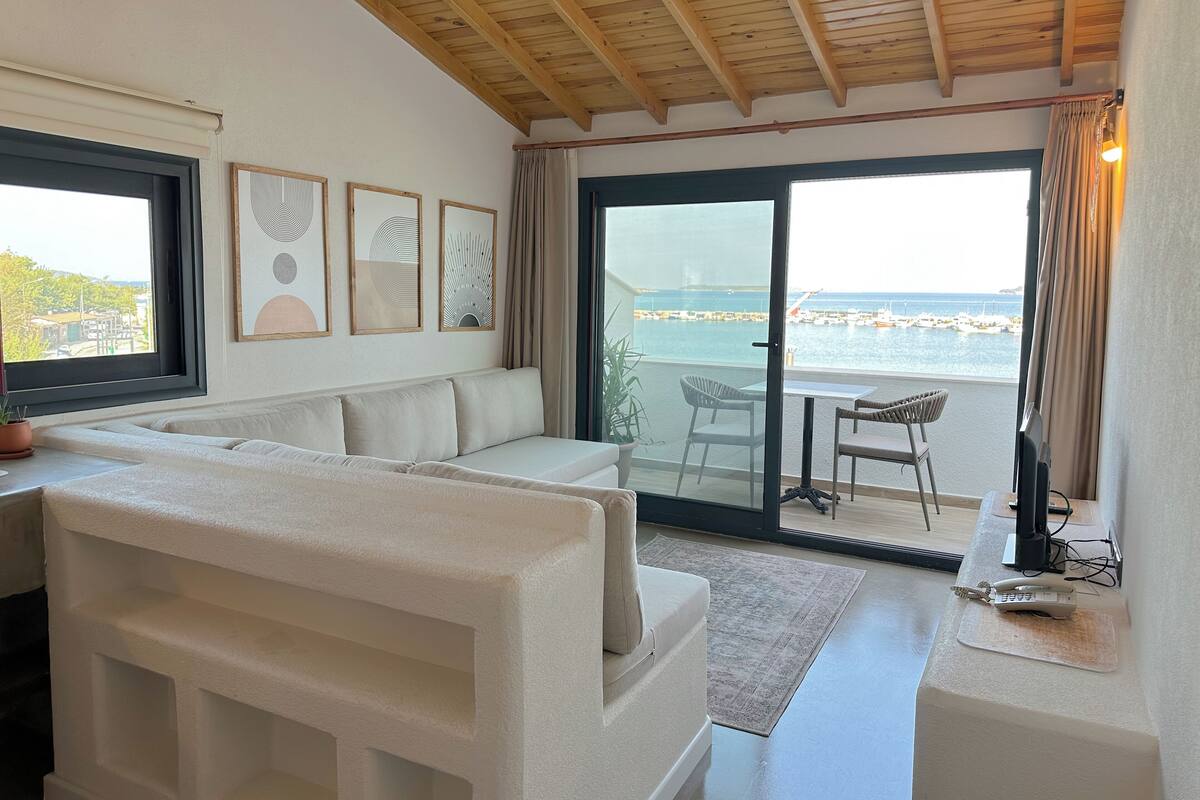 Uma sala de estar moderna com um sofá branco, uma mesa de jantar com cadeiras e vista para o mar através de grandes portas de vidro.