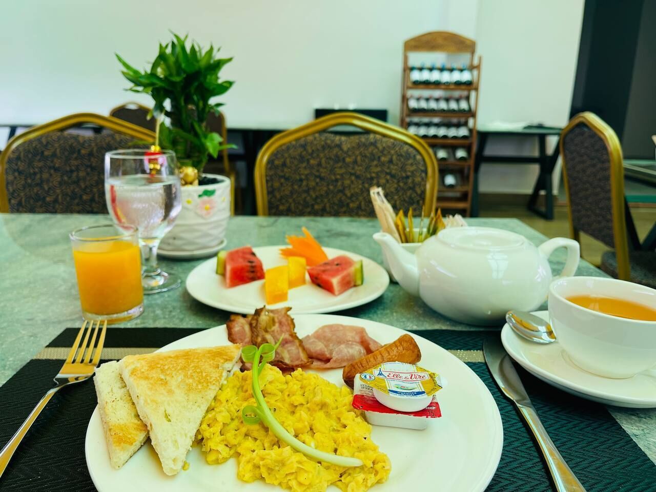 Disposição de café da manhã com torradas, ovos mexidos, bacon e frutas em um prato, um bule, uma xícara de chá e um copo de suco de laranja.