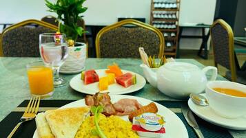 Disposição de café da manhã com torradas, ovos mexidos, bacon e frutas em um prato, um bule, uma xícara de chá e um copo de suco de laranja.