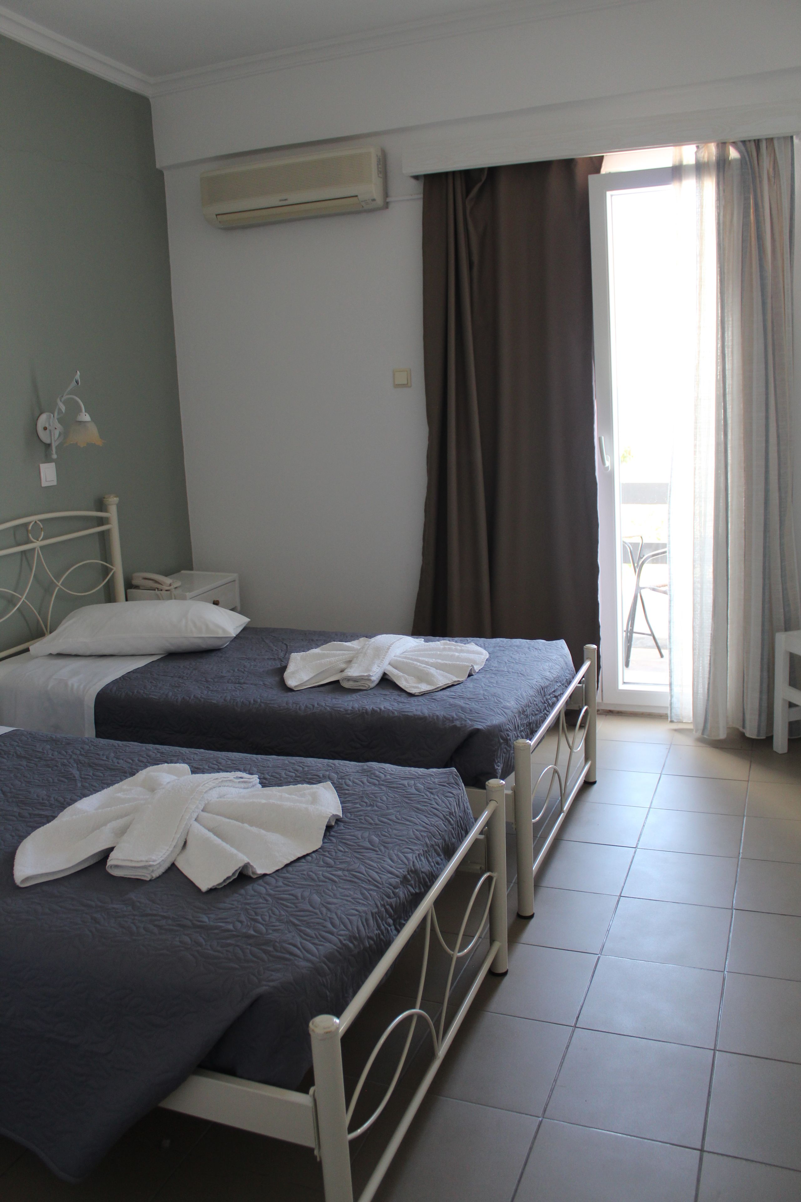 Quarto de hotel com duas camas, uma mesa de cabeceira, uma luminária e uma janela com cortinas.