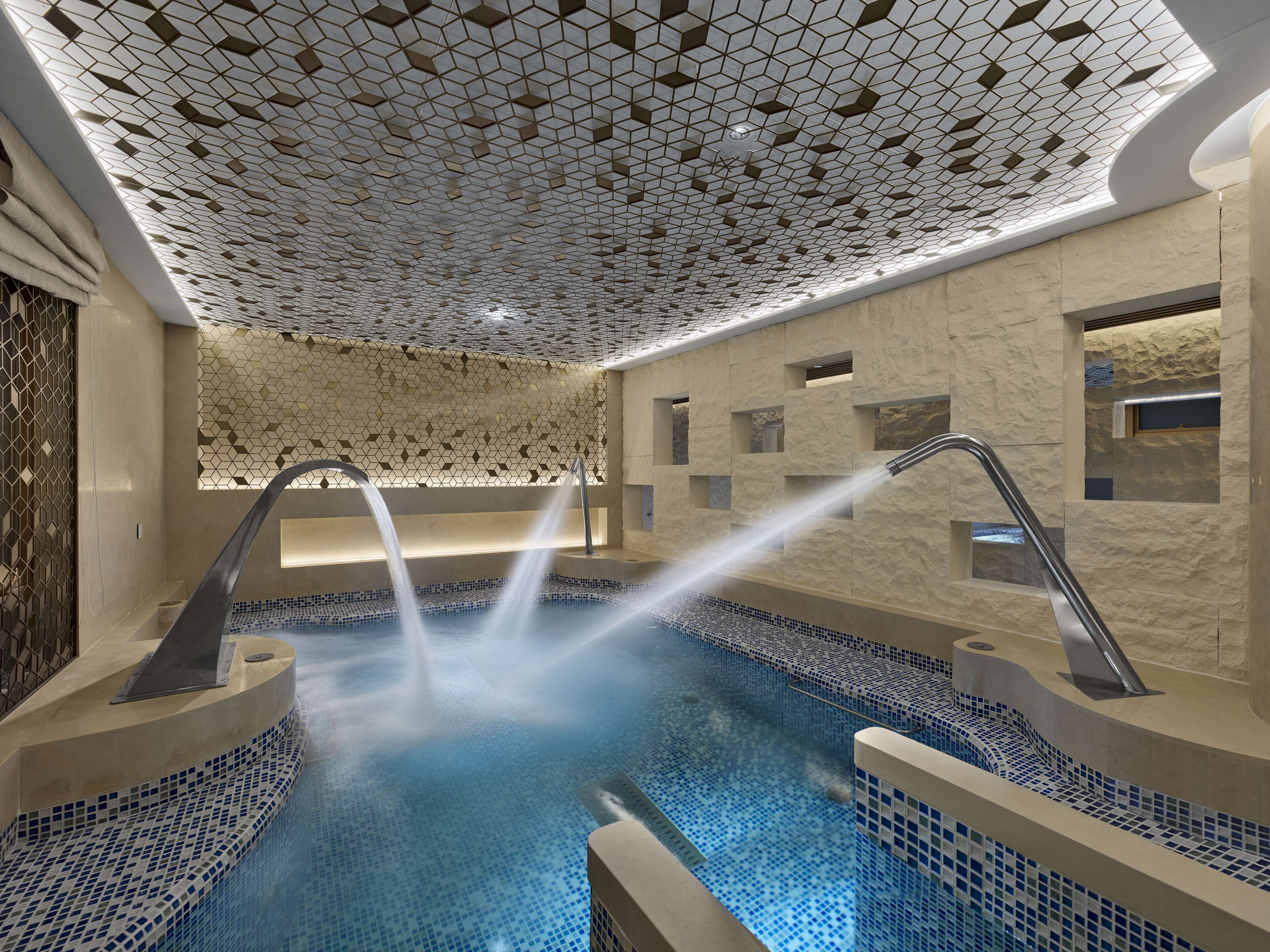 Piscina interior moderna com design geométrico no teto, jato de água e parede com textura de pedra.
