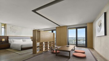 Quarto de hotel moderno com uma cama, uma escrivaninha, uma cadeira, uma mesa pequena e vista da cidade através de grandes janelas.