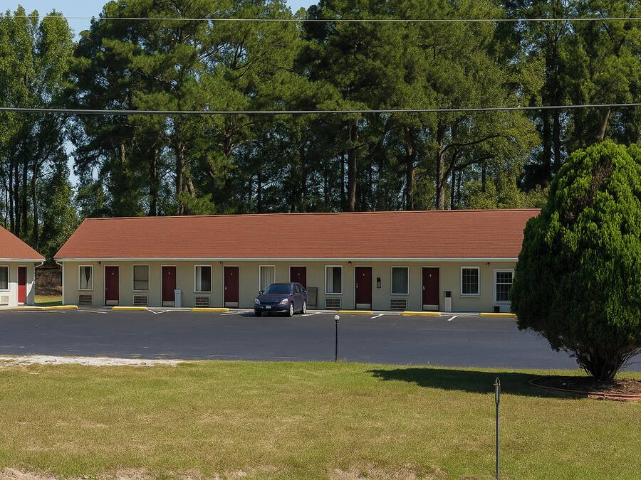 Um motel com telhado vermelho e várias salas, um estacionamento e uma grande árvore em primeiro plano.