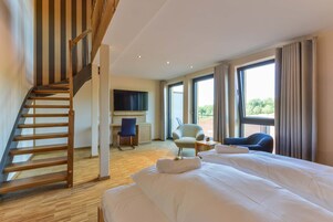 Quarto de hotel com escada de madeira, cama com roupa de cama branca, escrivaninha com cadeira e grandes janelas com vista.
