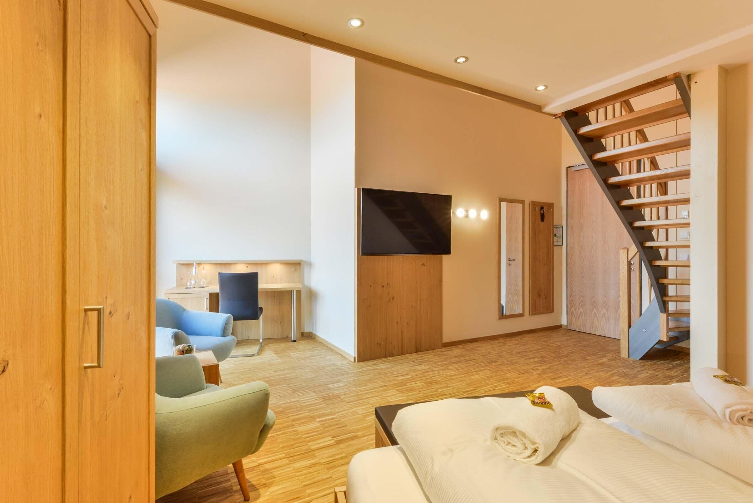 Quarto de hotel moderno com escada de madeira, televisão de tela plana e área de estar com sofá e poltronas.