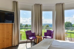 Quarto de hotel com uma cama grande, duas cadeiras roxas, uma mesa pequena e uma TV.