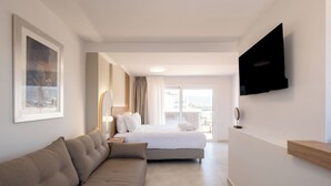 Quarto de hotel moderno com cama, sofá, TV fixada na parede e uma janela grande.