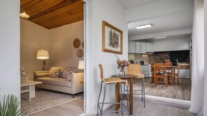 Uma sala de estar moderna com sofá, duas poltronas, mesa de centro e área de jantar com mesa e cadeiras. Uma cozinha é visível ao fundo.
