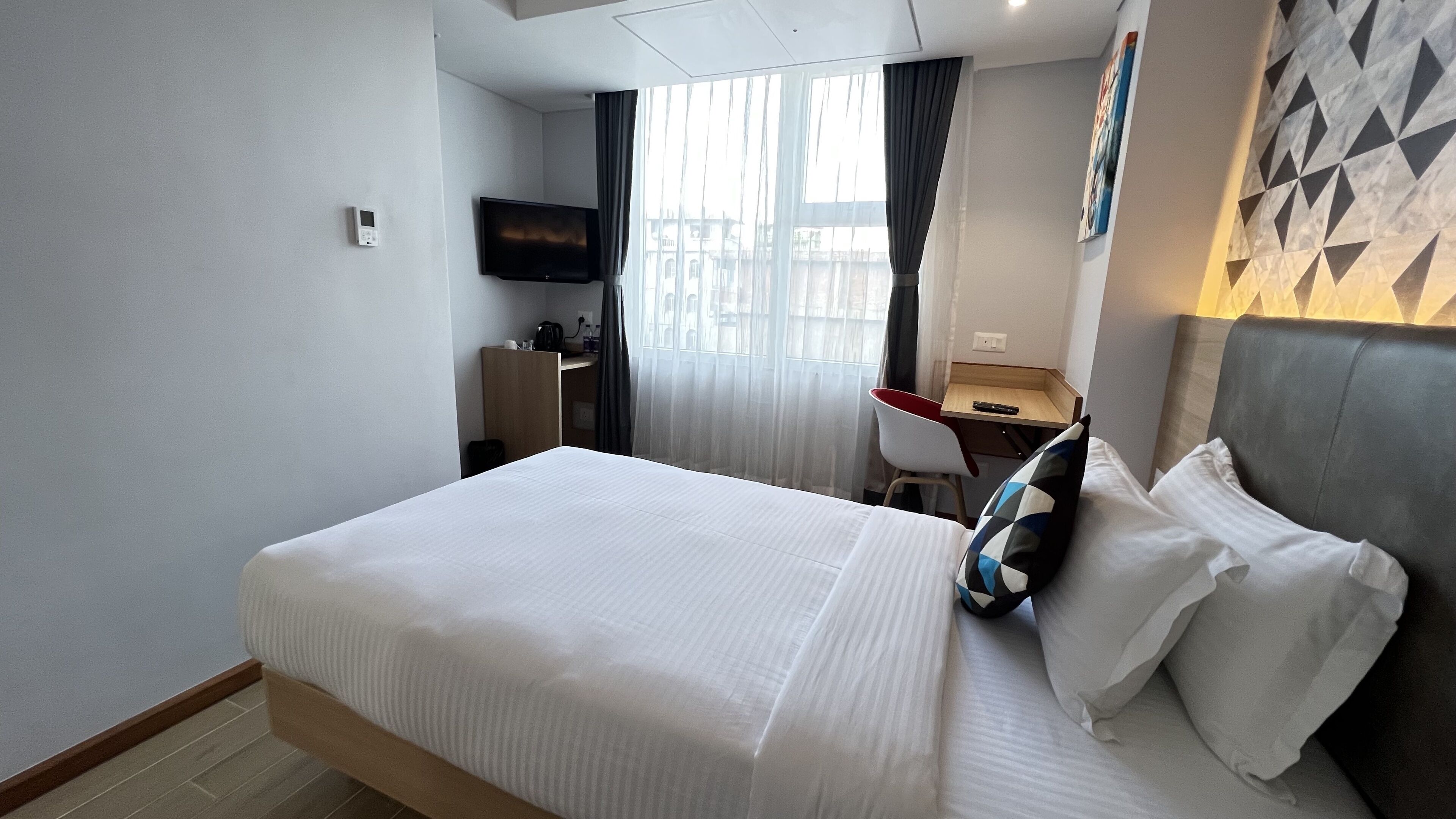 Quarto de hotel com cama, escrivaninha, cadeira, televisão e janela com cortinas.