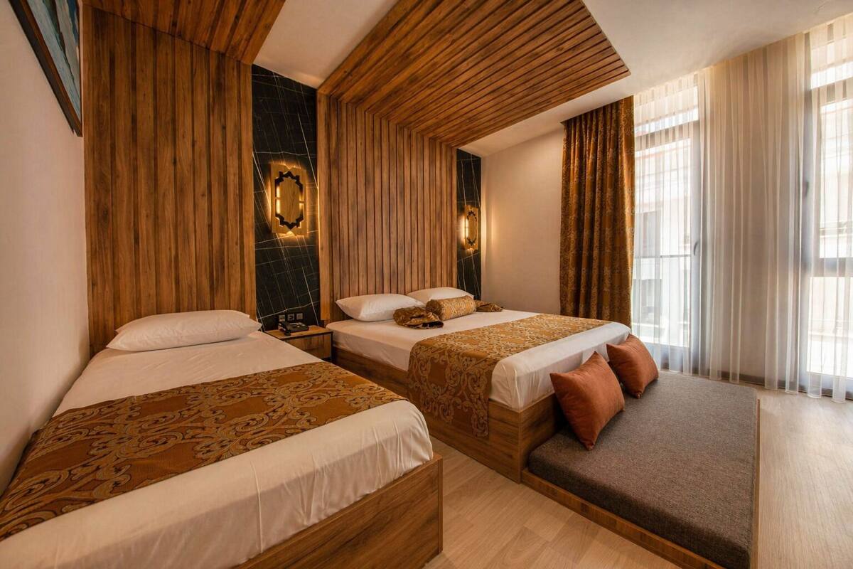 Quarto de hotel com duas camas, um sofá, painéis de madeira e uma janela grande com cortinas.